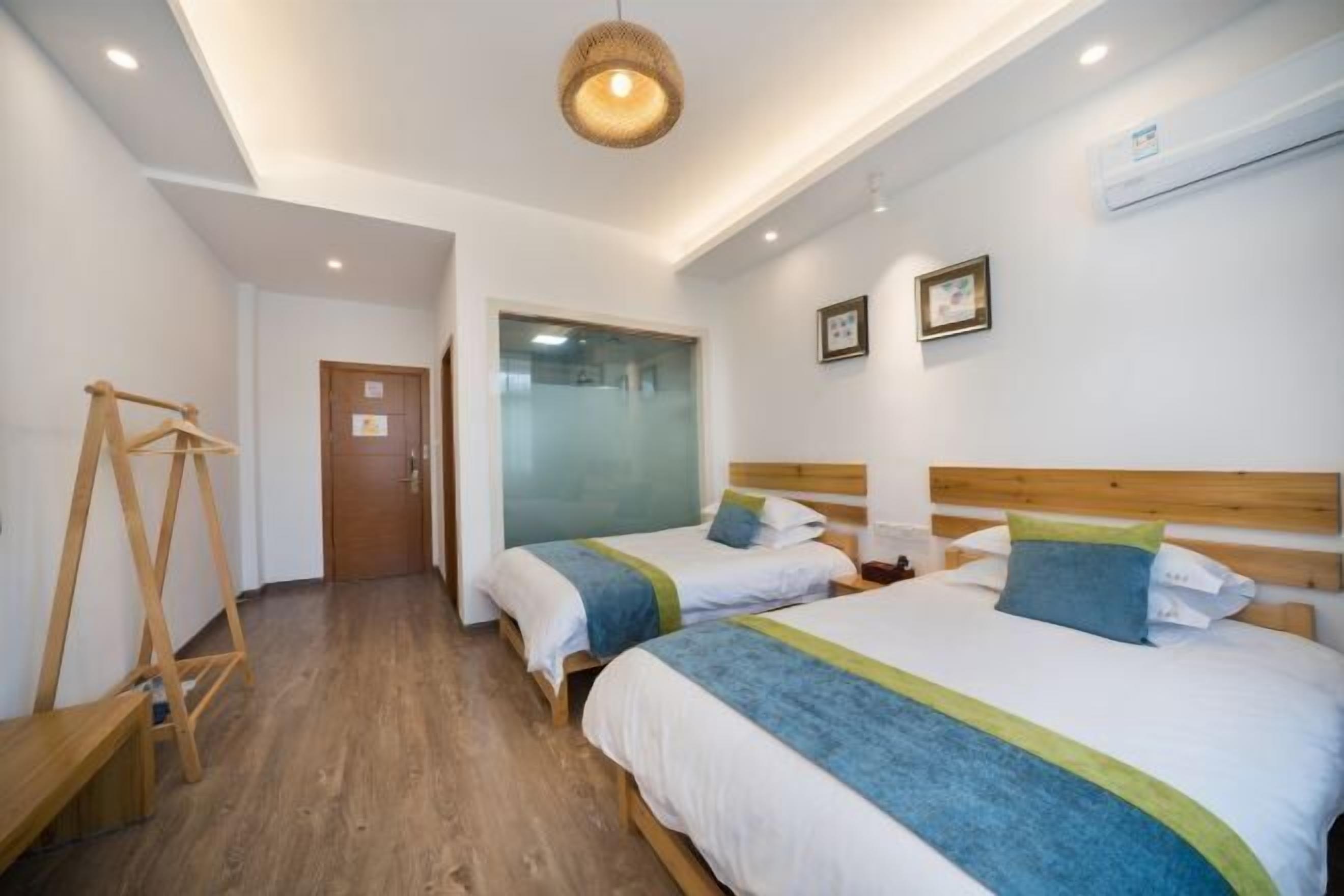 Quarto de hotel com duas camas, cabeceiras de madeira, um banco de madeira, um candeeiro pendente e uma porta que dá para outro quarto.