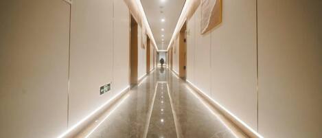 Corredor com piso lustroso, luminárias fixadas na parede e placa indicando saída de emergência.