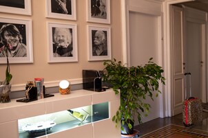 Uma área de recepção com fotografias a preto e branco emolduradas na parede, uma planta em vaso, uma máquina de café e uma mala.