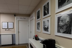 Um corredor com fotografias emolduradas na parede, uma porta branca com a inscrição "Pucci Filli" e um aparador branco com um objeto preto em cima.