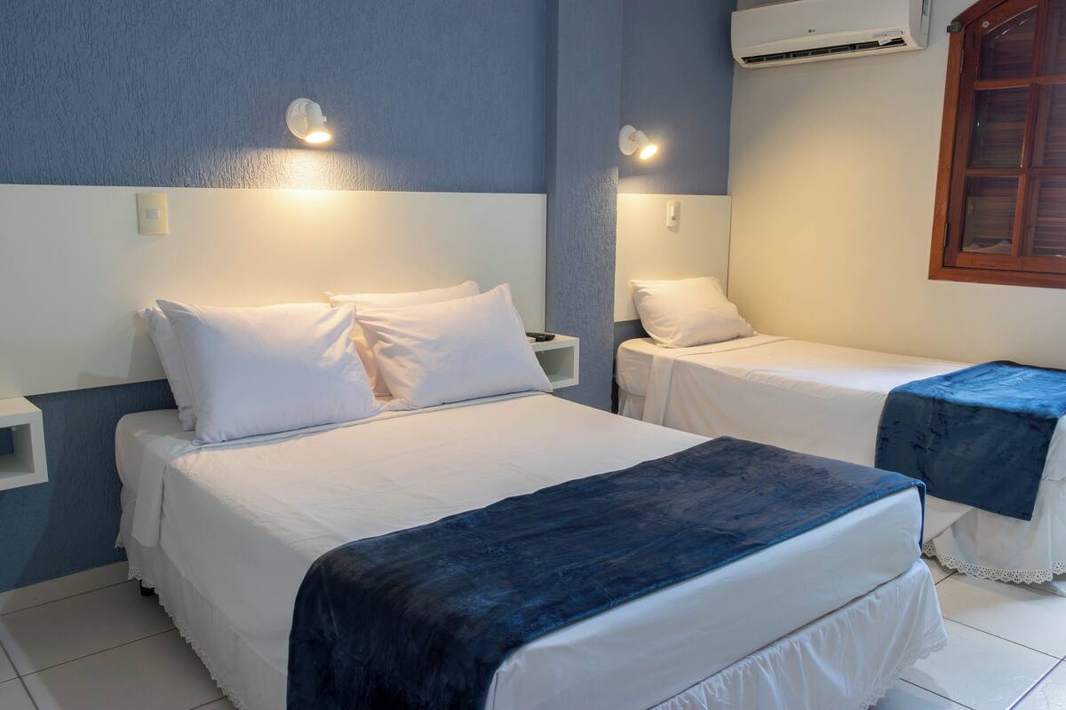 Quarto de hotel com duas camas, uma mesa de cabeceira, ar condicionado fixado na parede e uma janela com moldura de madeira.
