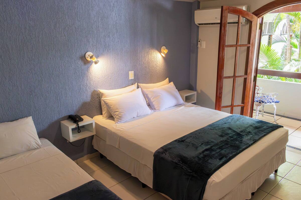 Quarto com cama, mesas de cabeceira, telefone e janela com vista para área verde.