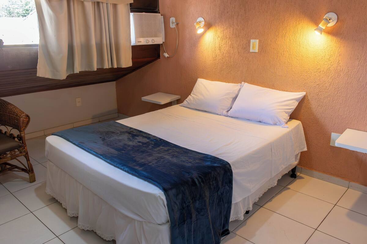 Cama bem arrumada com roupa de cama branca e um cobertor azul, uma pequena mesa de cabeceira, um aquecedor fixado na parede e um candeeiro também fixado na parede.