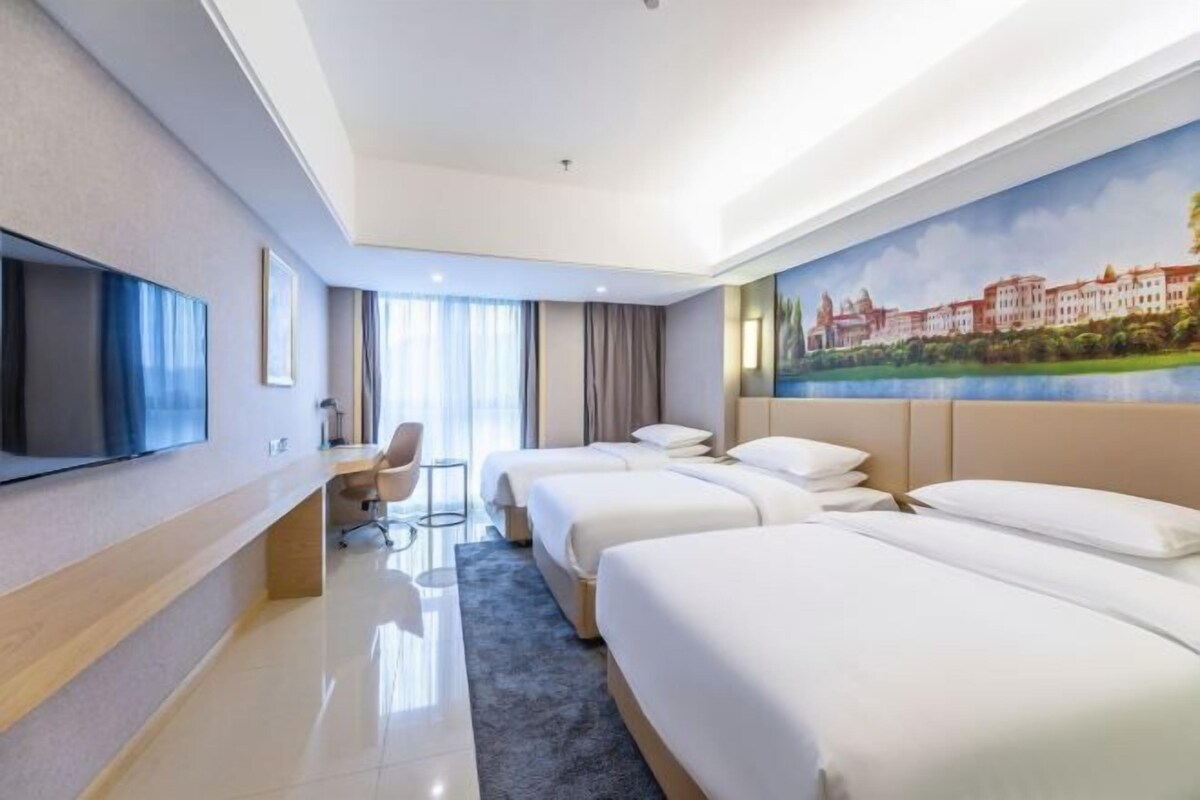 Quarto de hotel com três camas, uma escrivaninha, uma cadeira e uma televisão grande.