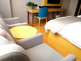Quarto de hotel com cama, escrivaninha com cadeira, mesa redonda pequena e cadeira azul.
