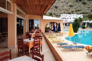 Uma área à beira da piscina com mesas e cadeiras, uma piscina e um edifício ao fundo.