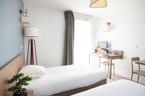 Quarto de hotel com duas camas, uma escrivaninha com cadeira, televisão, abajur e planta em vaso.
