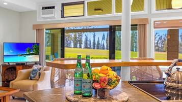Quarto de hotel moderno com uma TV exibindo uma praia tropical, um bar com garrafas e frutas, e vista para um campo de golfe.