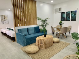 Quarto de hotel moderno com um sofá azul, área de jantar com mesa redonda e cadeiras, e uma grande planta em vaso.