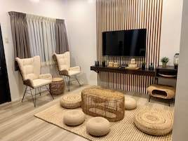 Uma sala de estar moderna com uma TV de tela plana, duas poltronas brancas acolchoadas, uma mesa de centro de madeira e pufes trançados sobre um tapete bege.