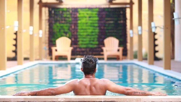 Um homem relaxa numa piscina, com cadeiras e uma parede verde ao fundo.