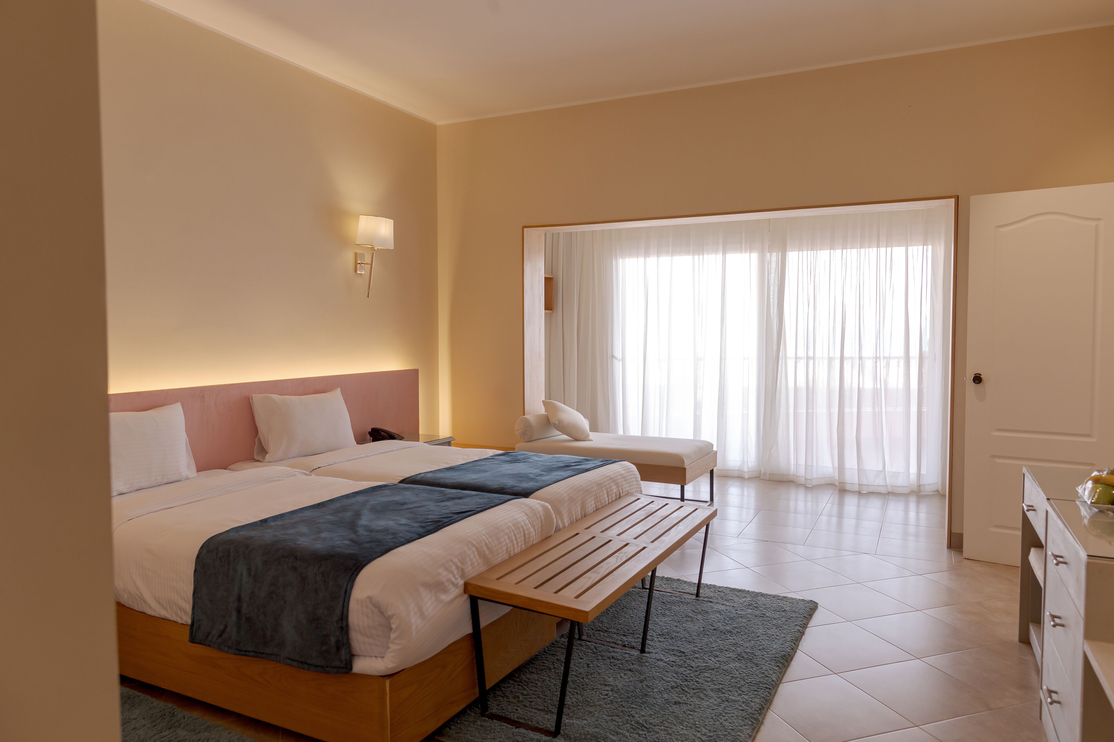 Quarto de hotel com cama de casal, mesa de cabeceira, um banco, escrivaninha e uma janela grande com cortinas translúcidas.