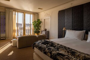 Quarto de hotel com uma cama grande, um sofá, uma planta em vaso e vista para a cidade.