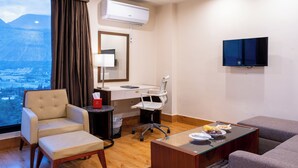 Quarto de hotel com vista para uma montanha, uma secretária com cadeira, um sofá, uma televisão e uma mesa de jantar com um prato de comida.