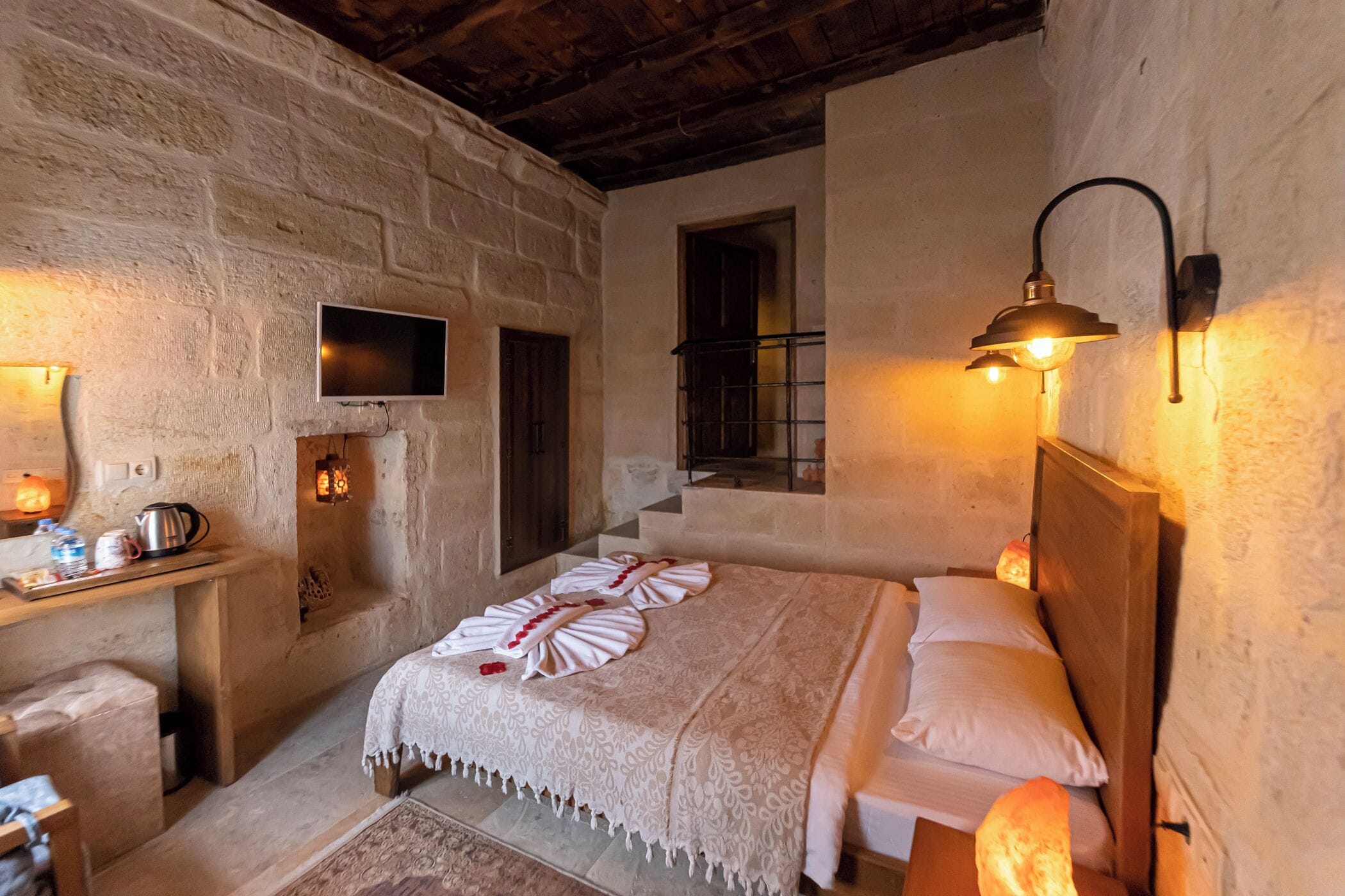 Quarto aconchegante com parede de pedra, cama de madeira, televisão, escrivaninha com chaleira e escada.