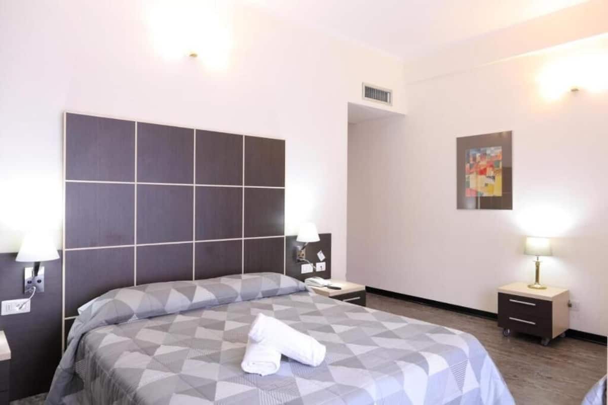 Quarto de hotel com uma cama grande, duas mesas de cabeceira com luminárias, uma obra de arte na parede e uma porta que dá para outro quarto.