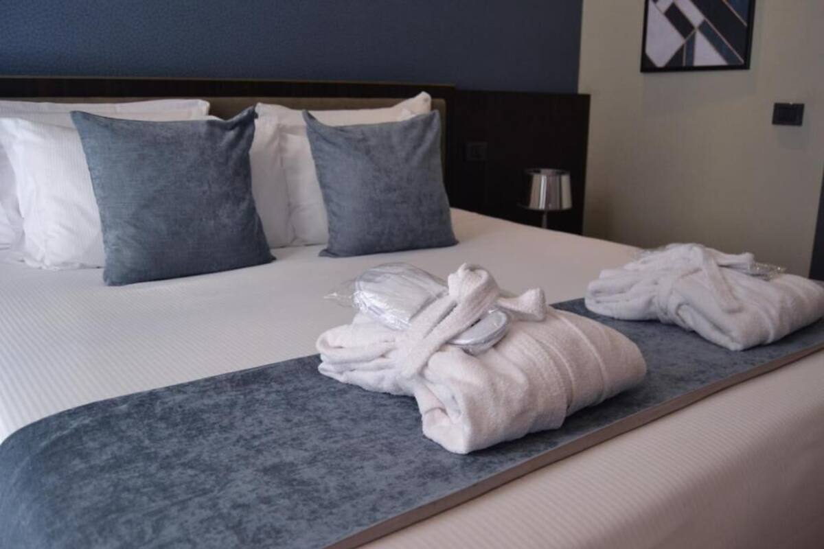 Cama bem arrumada com almofadas brancas e cinzentas, cabeceira cinza e duas toalhas sobre uma mesinha de cabeceira cinza.