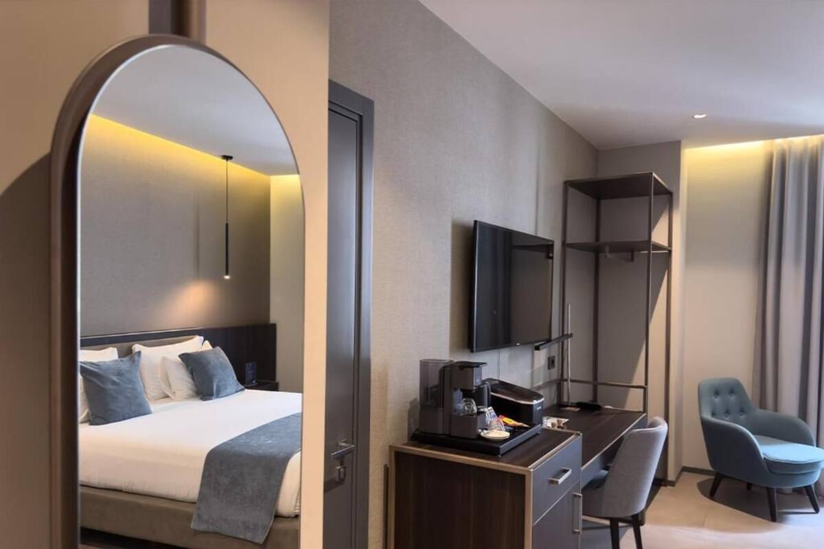Quarto de hotel moderno com uma cama, uma escrivaninha com cafeteira, uma televisão e uma cadeira.