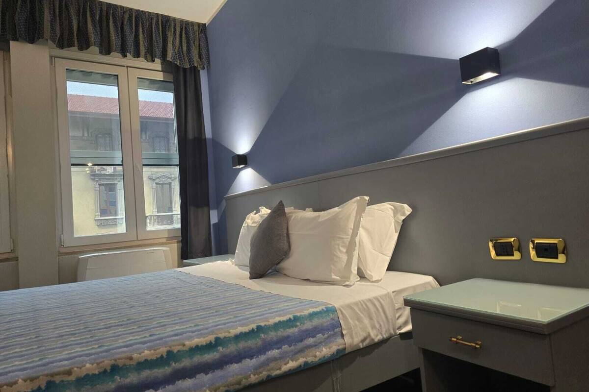 Quarto com cama, mesa de cabeceira e janela com cortinas.