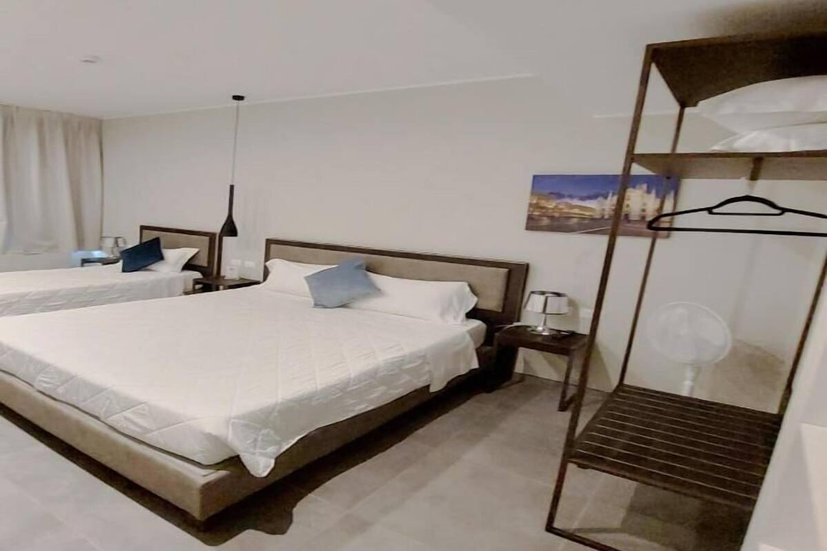 Quarto de hotel com duas camas, uma mesa de cabeceira com abajur, um cabideiro e um quadro na parede.
