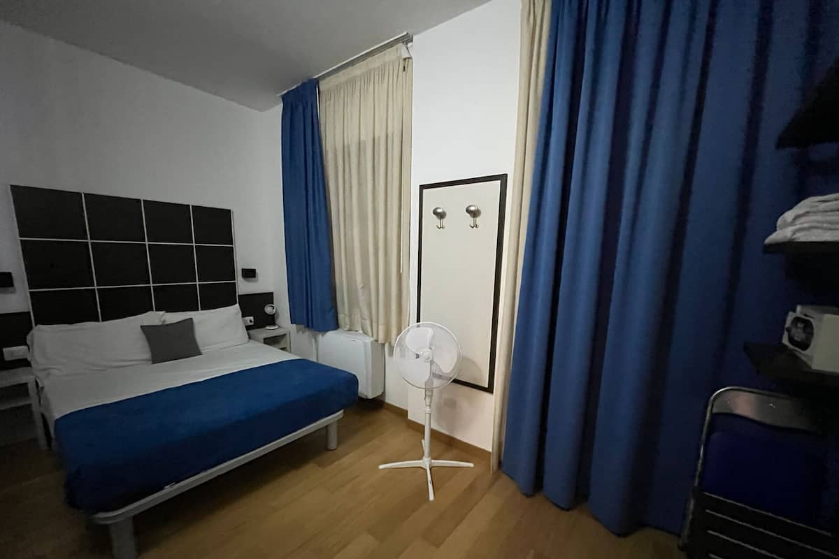 Quarto de hotel com cama, mesa de cabeceira, luminária e ventilador.