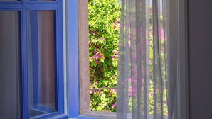 Janela com moldura azul e cortina branca, dando para um jardim com flores cor-de-rosa.