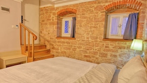 Quarto com parede de pedra, duas janelas com cortinas, uma cama de madeira e uma mesinha de cabeceira.