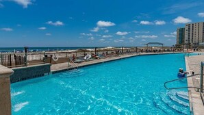 Uma piscina com água azul cristalina, rodeada por espreguiçadeiras e guarda-sóis, com vista para a praia e o oceano ao fundo.