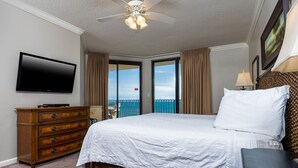 Quarto de hotel com cama, televisão, cômoda, ventilador de teto e vista para o oceano através de uma porta deslizante.