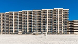 Um hotel alto com vários andares e varandas, localizado em uma praia arenosa sob um céu azul límpido.