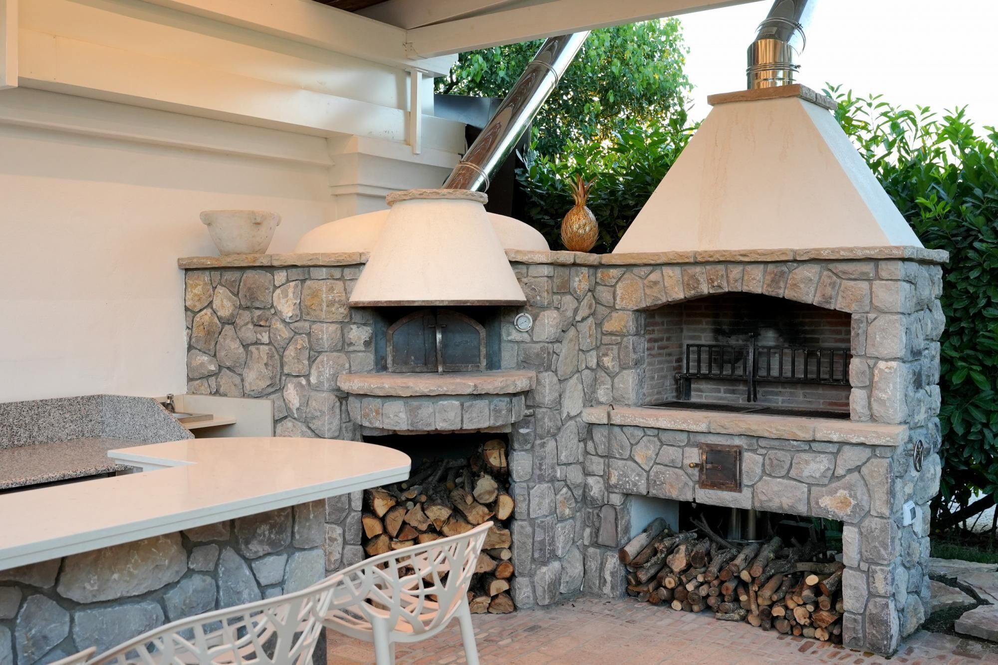 Forno de pizza de pedra ao ar livre com chaminé, área de estar com mesa e cadeiras, e uma pilha de lenha por perto.