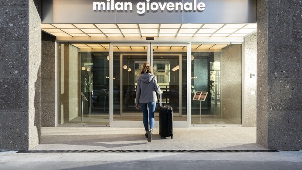 A building entrance with the sign "milan giovenale" and a person with a suitcase.