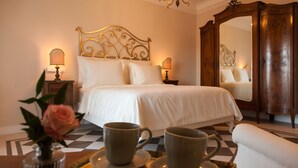 Quarto de hotel com cama, abajures, espelho, mesa de centro com xícaras e um vaso com uma flor.