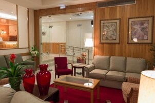 Um saguão de hotel com carpete vermelho, uma poltrona vermelha, um sofá bege e uma mesa de centro de vidro. Há quadros nas paredes e um abajur sobre uma mesa lateral.