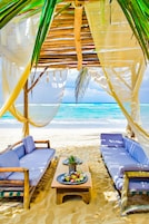 Uma cabana de praia com duas áreas de assento acolchoadas, uma mesa com uma travessa de frutas e vista para o oceano.