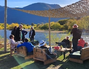 Um grupo de pessoas sentadas em bancos e mesas de madeira à beira de um lago, com uma montanha ao fundo.