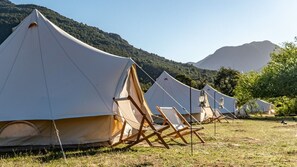 Fileira de grandes tendas brancas com cadeiras de madeira em frente, situada numa área gramada com montanhas ao fundo.