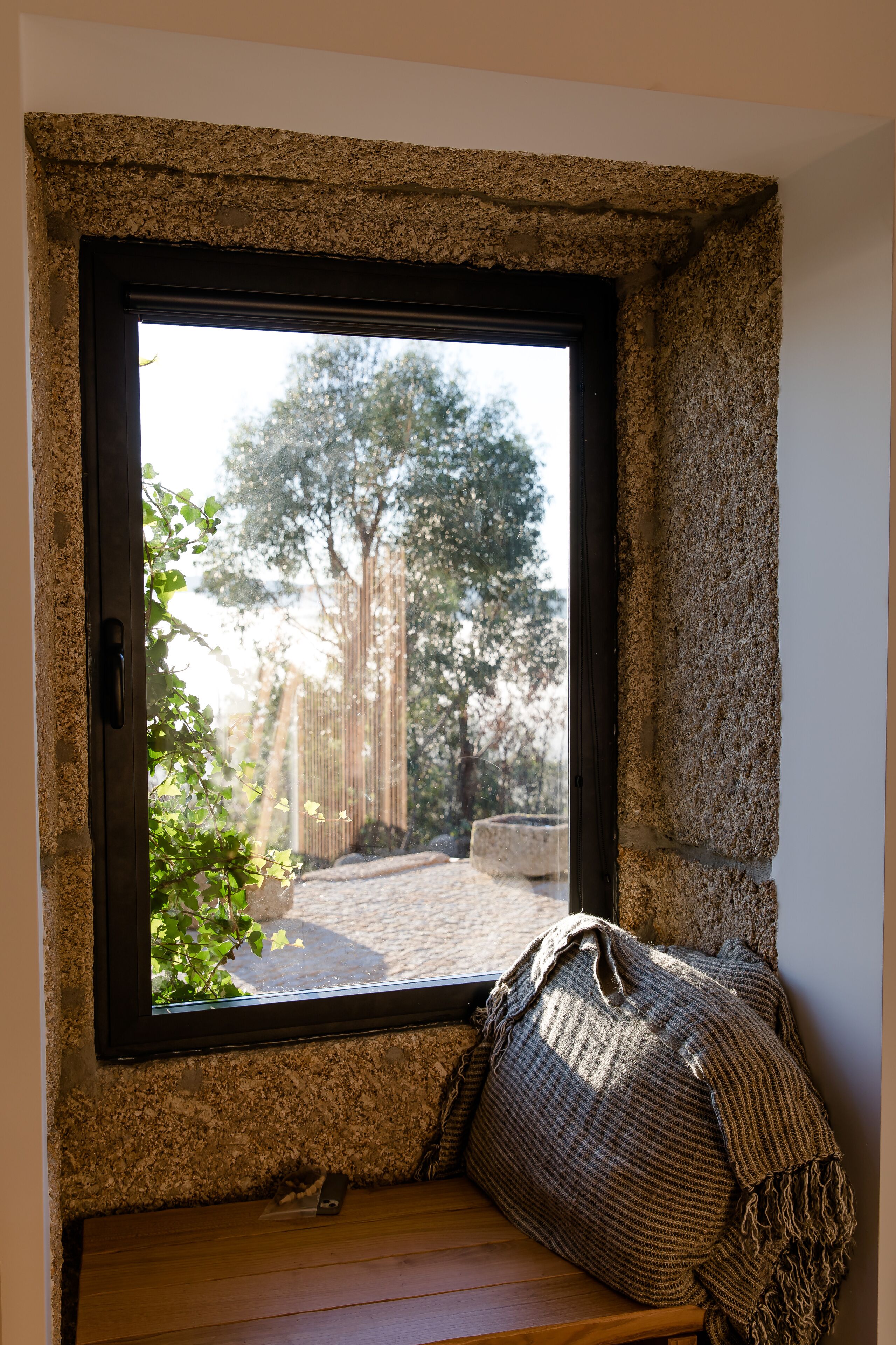 Janela com moldura de pedra, vista para árvores e um banco com cobertor.