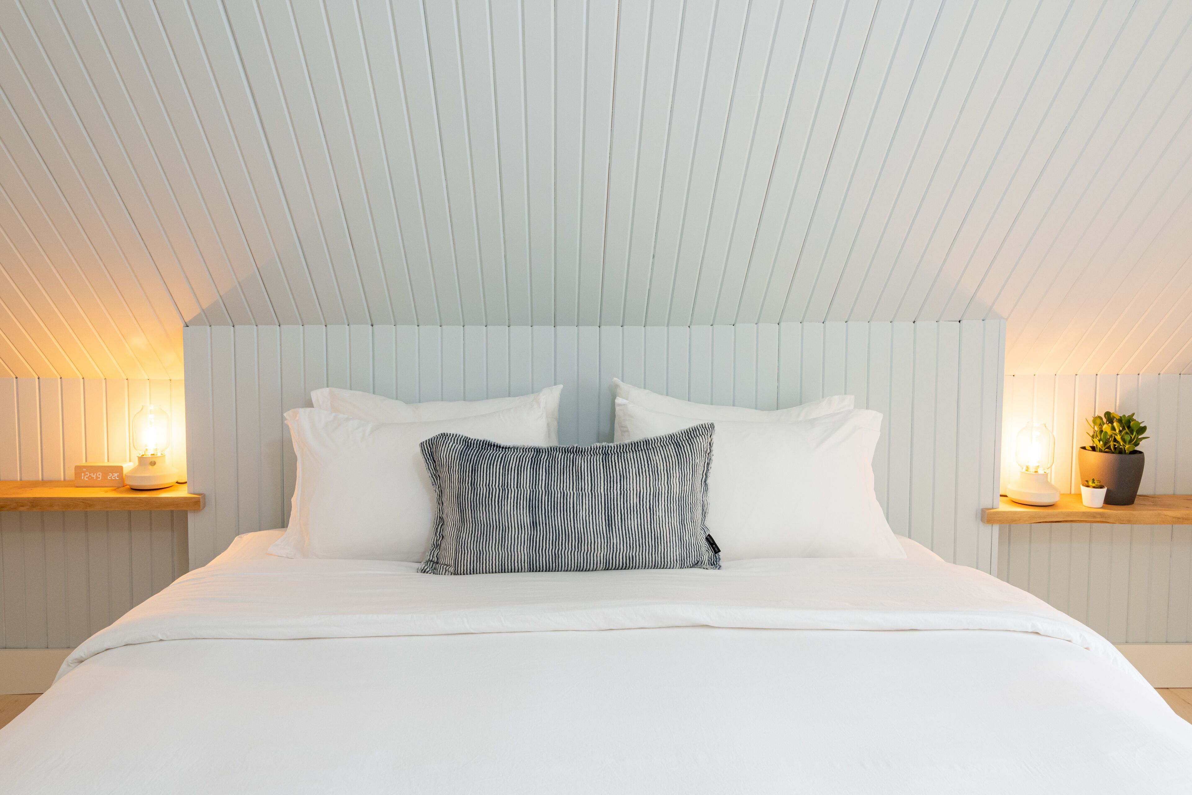 Quarto com teto inclinado, cama com roupa de cama branca, duas mesas de cabeceira com luminárias e um vaso de plantas em uma das mesas.