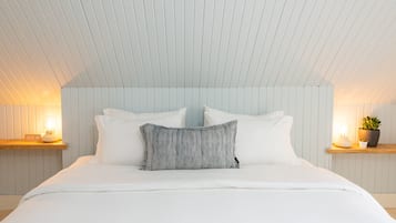 Quarto com teto inclinado, cama com roupa de cama branca, duas mesas de cabeceira com luminárias e um vaso de plantas em uma das mesas.