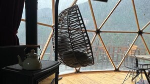 Um quarto aconchegante com piso de madeira, uma cadeira de balanço preta, uma chaleira branca em um fogão e uma grande janela com vista para uma área arborizada.