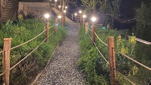 Um caminho noturno com piso de cascalho, ladeado por estacas e cordas de madeira, conduzindo a um edifício com fachada de pedra e escadaria.