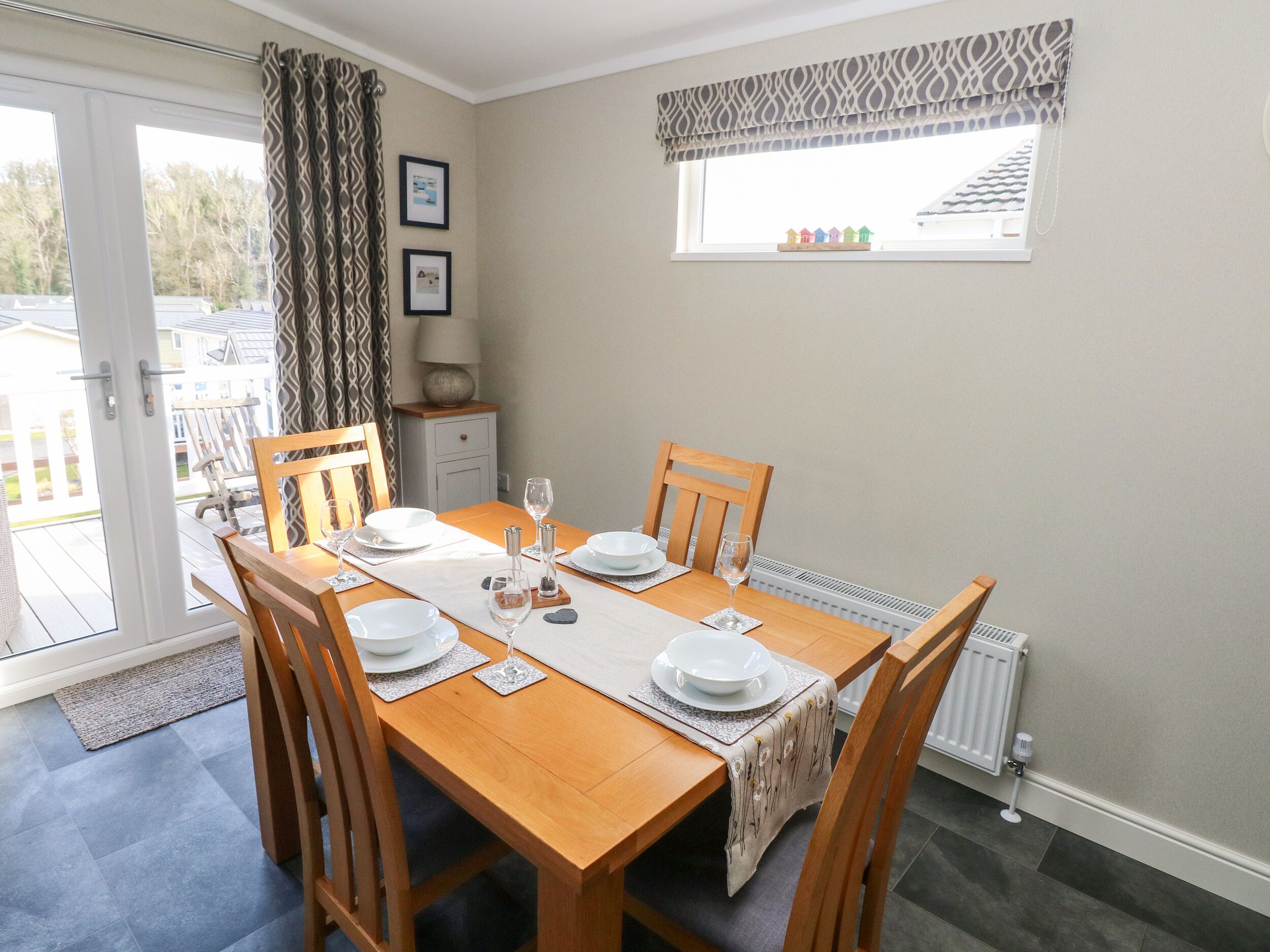 Uma área de refeições com mesa de madeira posta para quatro pessoas, cadeiras, janela com cortinas estampadas e vista para casas lá fora.