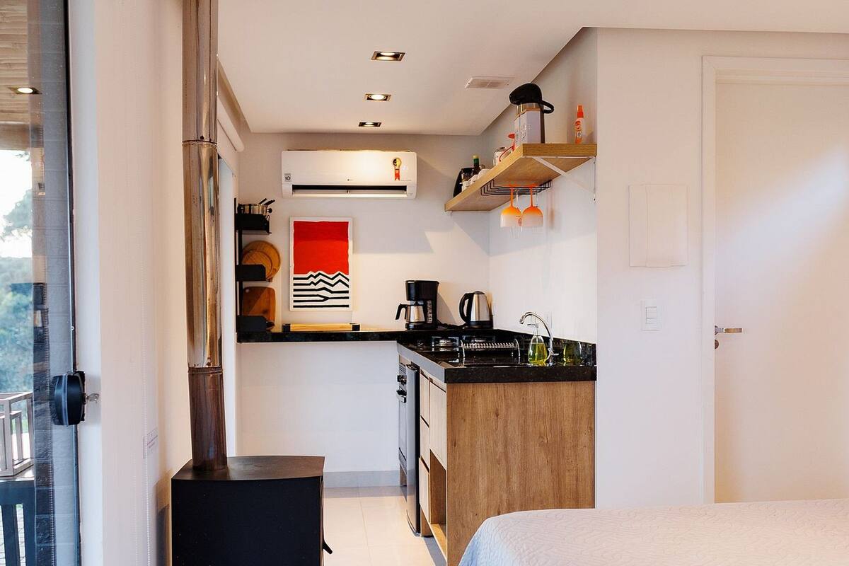 Quarto moderno com cama grande, um banquinho preto pequeno e uma cozinha com fogão e armários.