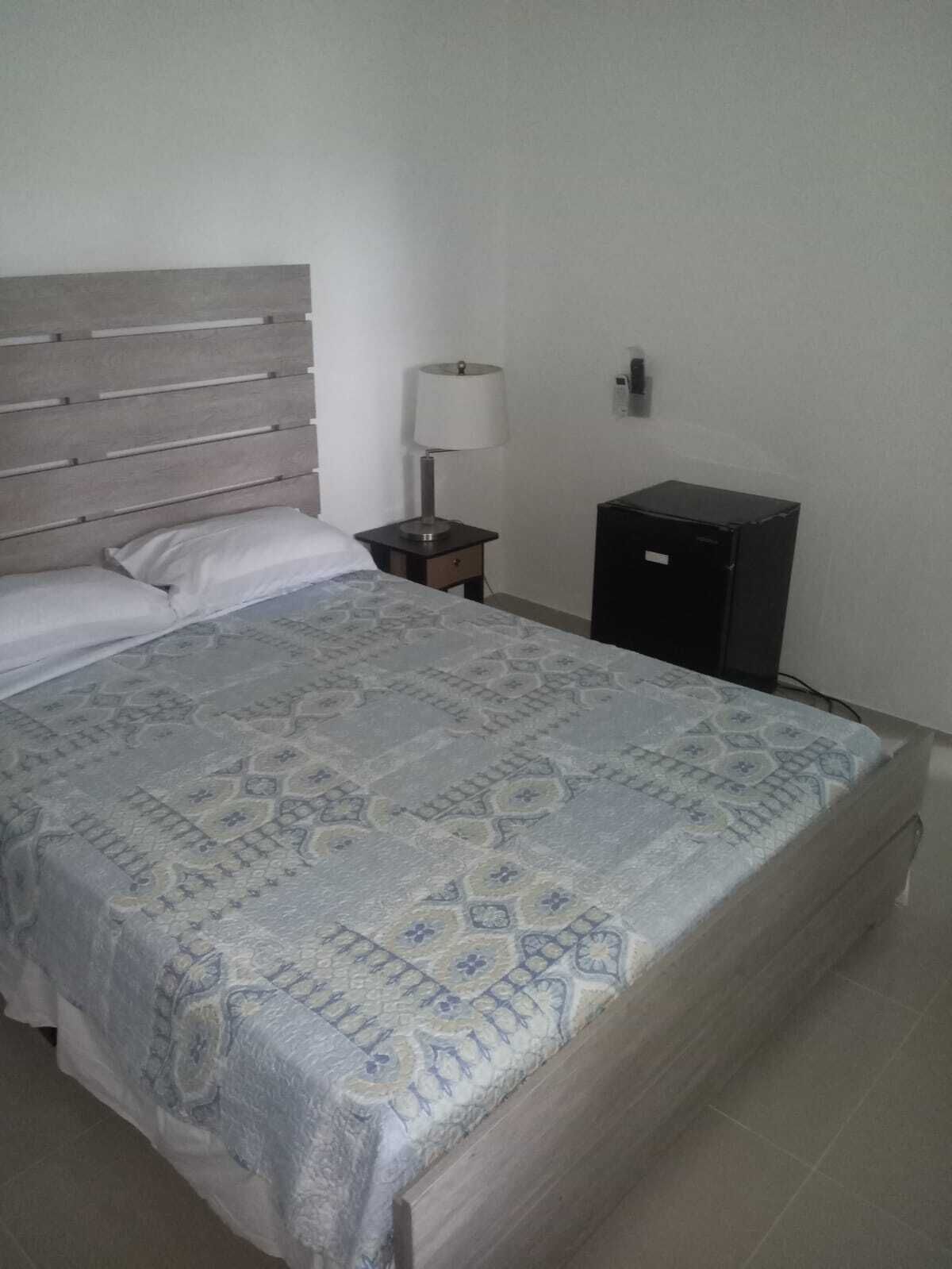 Quarto com cama, mesa de cabeceira, abajur e um pequeno armário preto.
