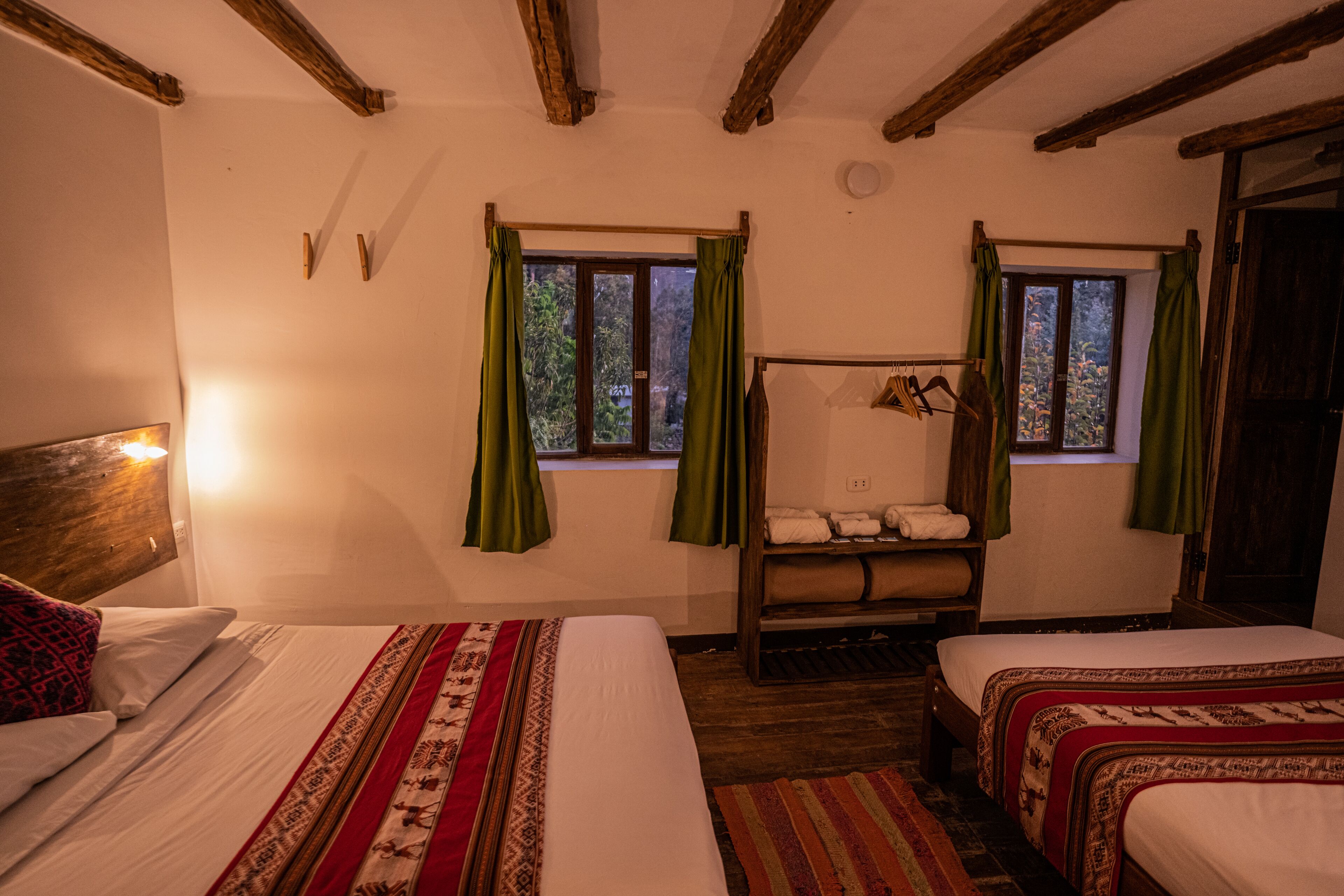 Quarto com duas camas, vigas de madeira no teto, uma janela com cortinas e uma estante com toalhas.