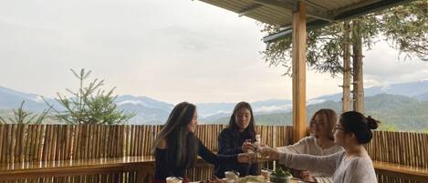Um grupo de pessoas sentadas em uma mesa de madeira, com vista para montanhas ao fundo.