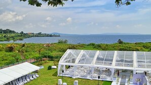 Uma estrutura em estilo de estufa com teto de vidro, rodeada por mesas e cadeiras brancas, com vista para um lago e vegetação.