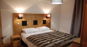 Quarto de hotel com cabeceira de madeira, duas luminárias de apoio, cama com roupa de cama branca e manta de pele.