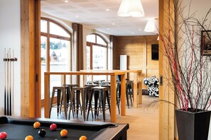 Uma mesa de bilhar com bolas, cadeiras de madeira, um bar e grandes janelas.